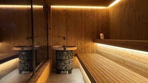 Sala de sauna com paredes e bancos de madeira, duas bacias de pedra e uma divisória de vidro.