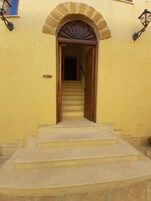 Uma escadaria leva a uma porta com arco de pedra, um pequeno letreiro na parede e duas luminárias fixadas na parede.