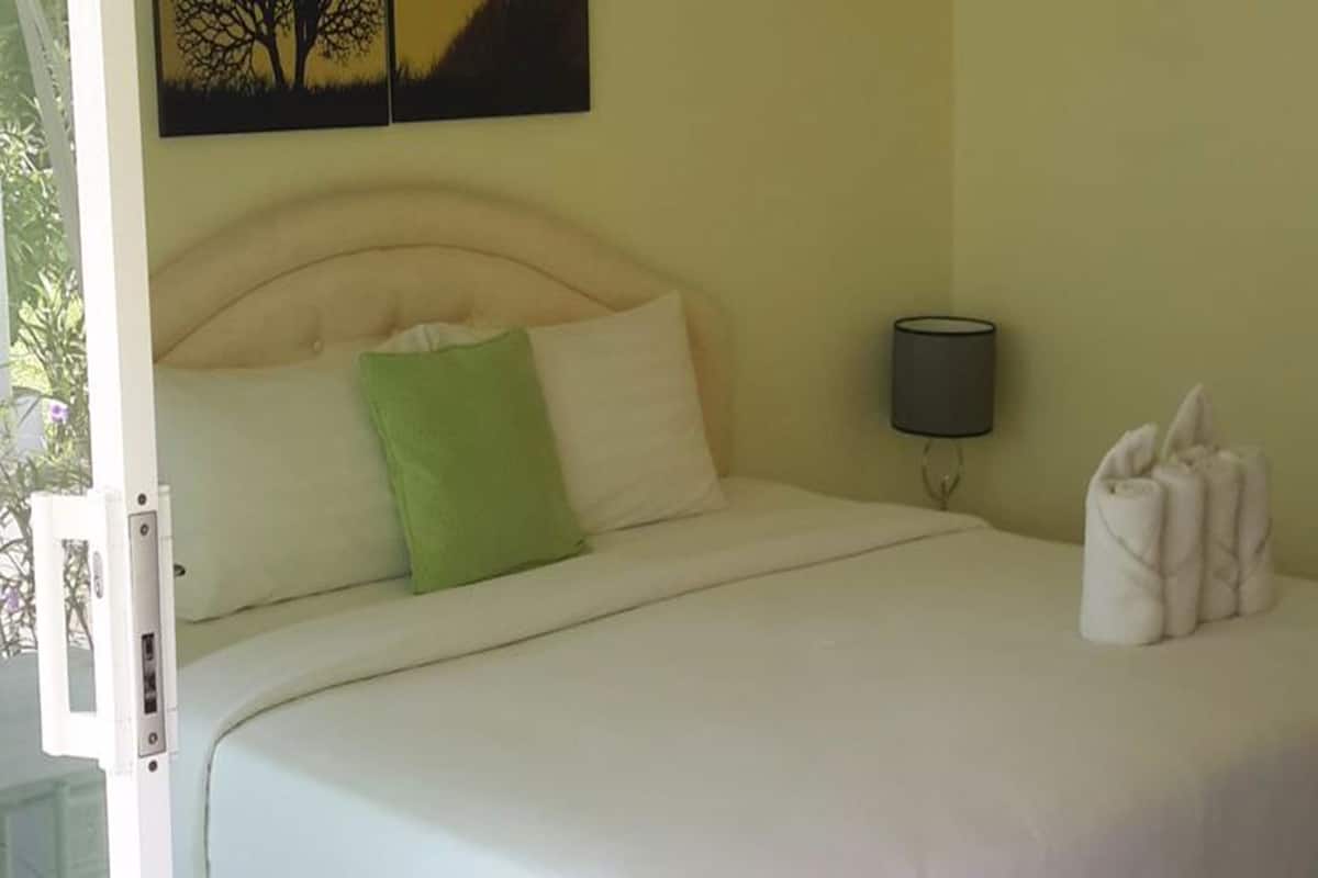 Quarto de hotel com cama, travesseiro verde, travesseiro branco, luminária de parede e um porta-toalhas com toalhas.