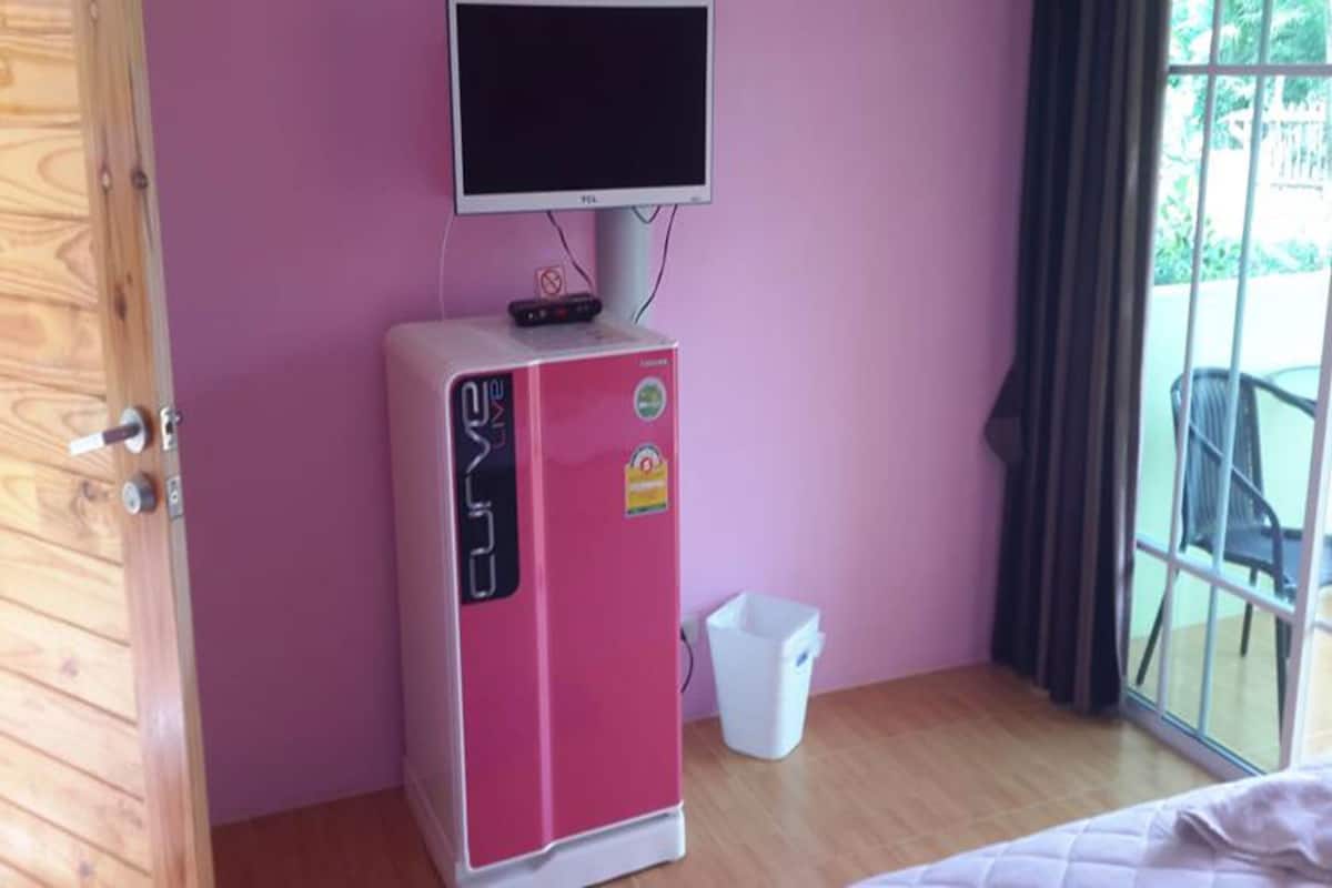 Quarto com parede rosa, cama, televisão sobre suporte, lixeira branca e janela com cortinas.