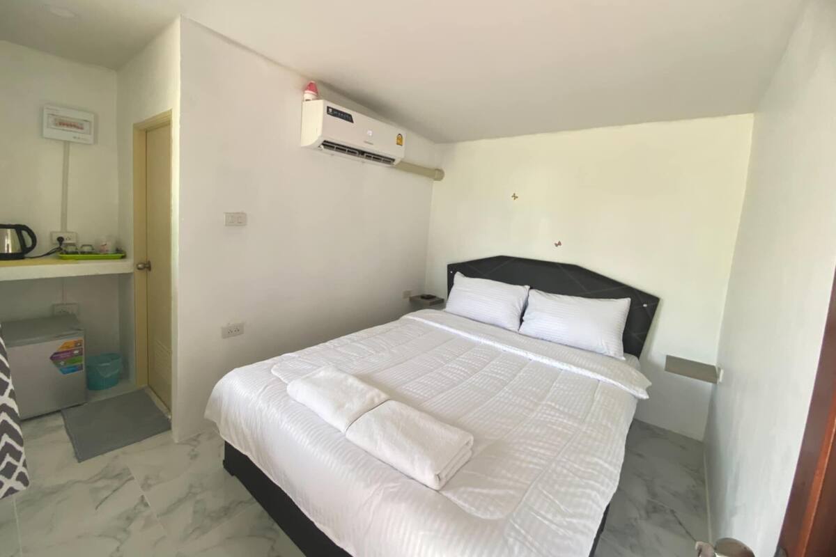 Quarto com cama, mesa de cabeceira, ar condicionado fixado na parede, prateleira com chaleira e lixeira pequena.