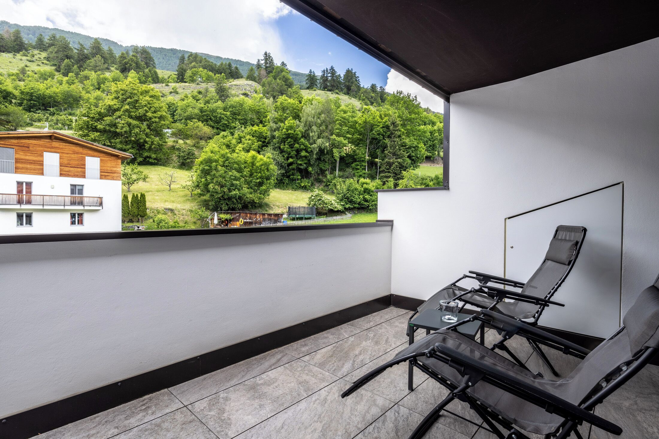 Uma varanda com vista para uma encosta verde, um edifício branco e uma mesa com duas cadeiras.