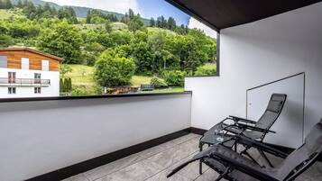 Uma varanda com vista para uma encosta verde, um edifício branco e uma mesa com duas cadeiras.