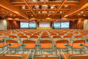 Sala de conferências com fileiras de cadeiras laranja, palco com tela e vigas de madeira no teto.