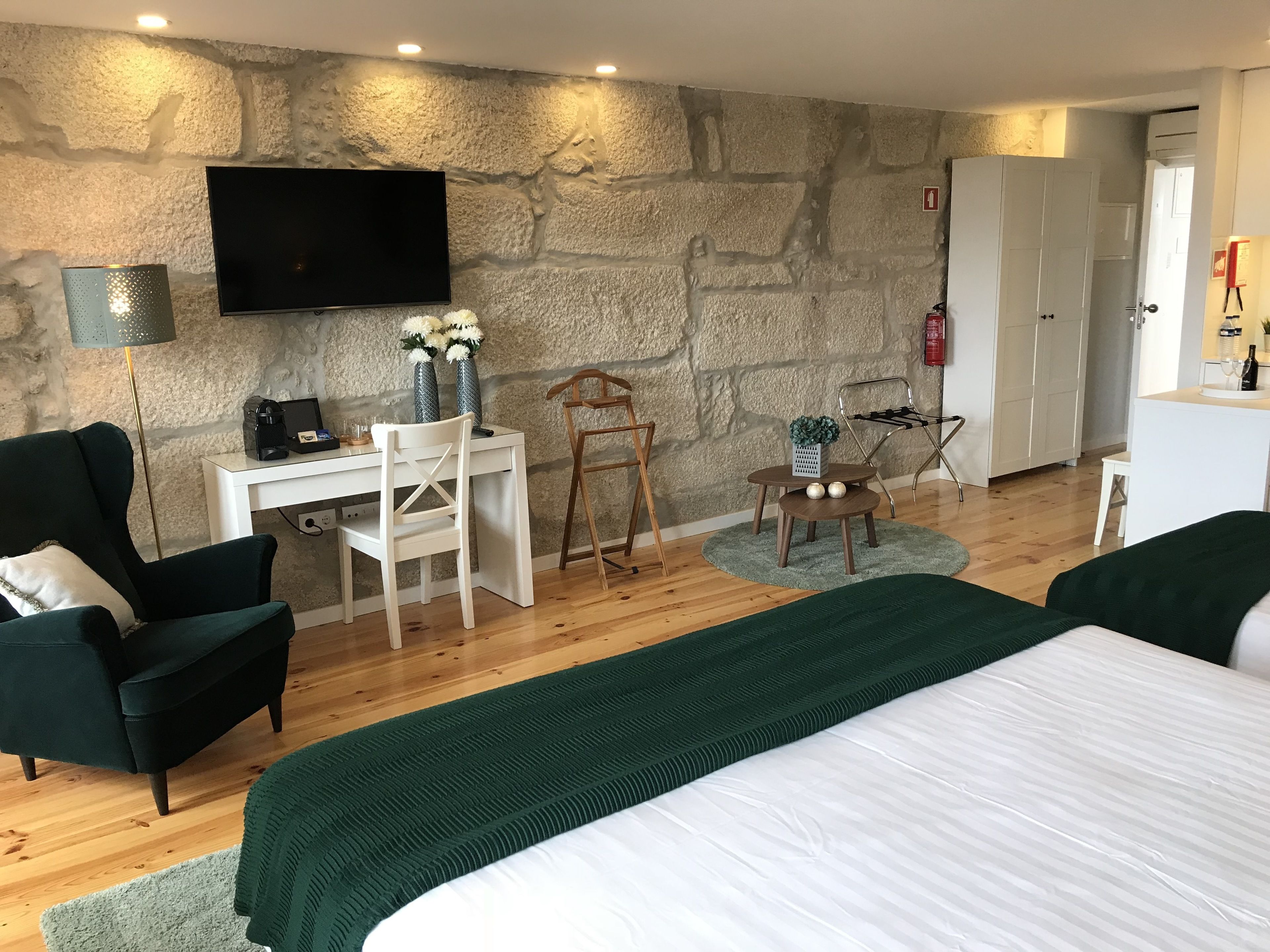 Quarto moderno com parede de pedra, televisão de tela plana, escrivaninha com cadeira, mesinha com objetos decorativos, guarda-roupa e cama com roupa de cama verde.