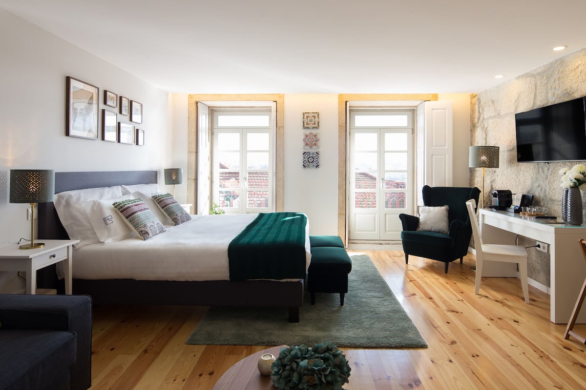 Quarto moderno com cama, mesas de cabeceira, escrivaninha, cadeira, televisão e vista pela porta.