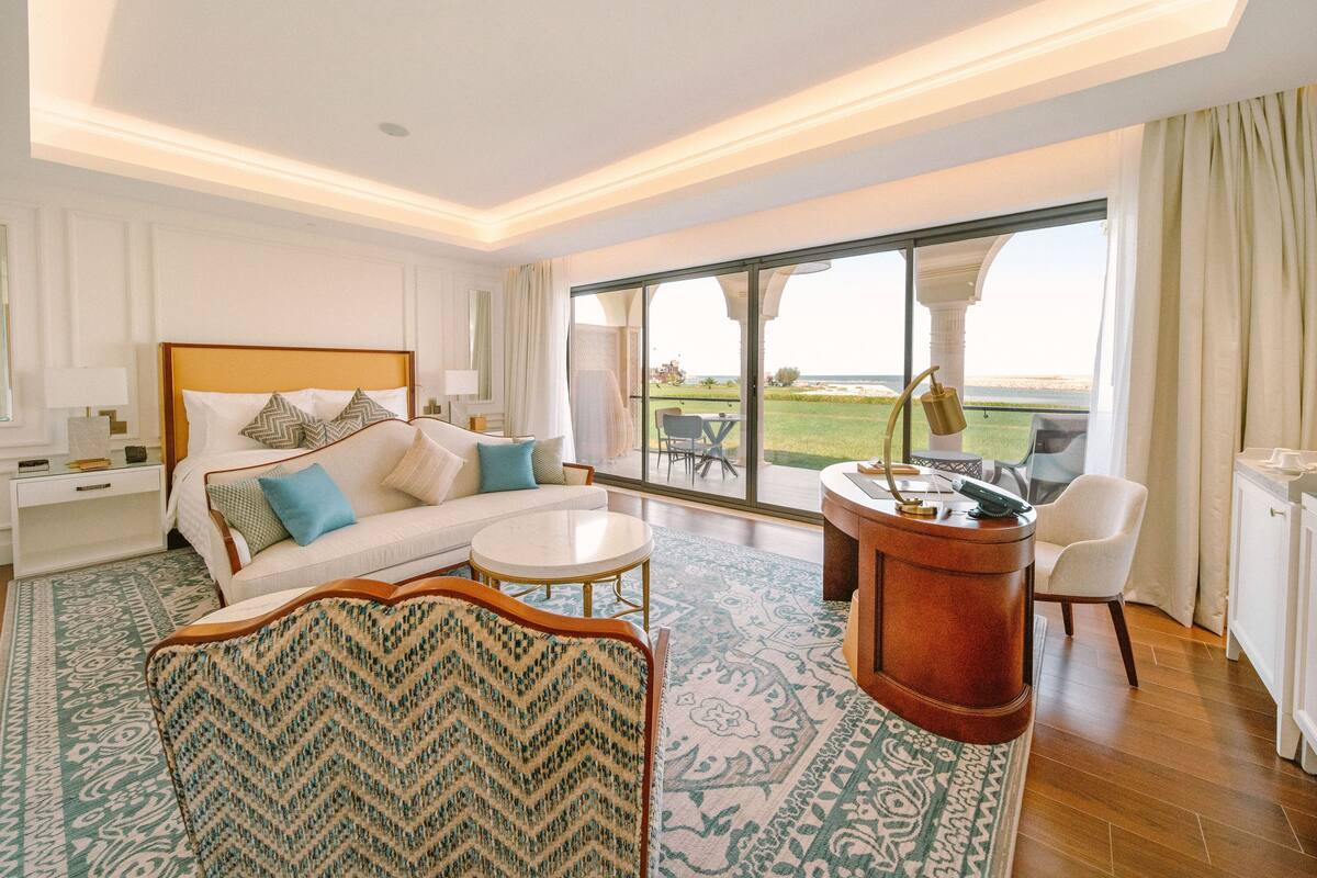 Quarto de hotel com uma cama grande, área de estar com mesinha de centro e vista para a praia através de grandes janelas.