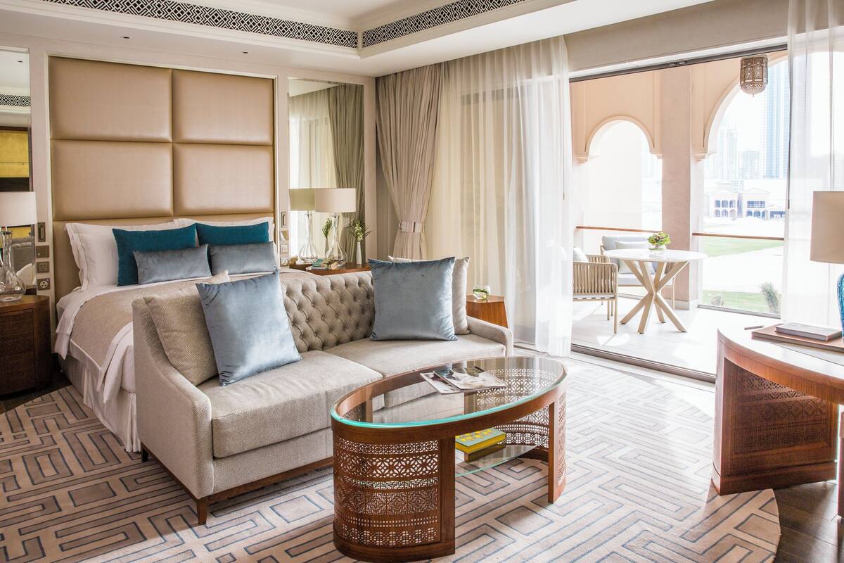Quarto de hotel com cama, sofá, mesa de centro de vidro e varanda com mesa e cadeiras.
