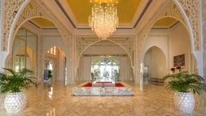 Um lobby luxuoso com piso de mármore, arcos elaborados, um lustre central e plantas em vasos.