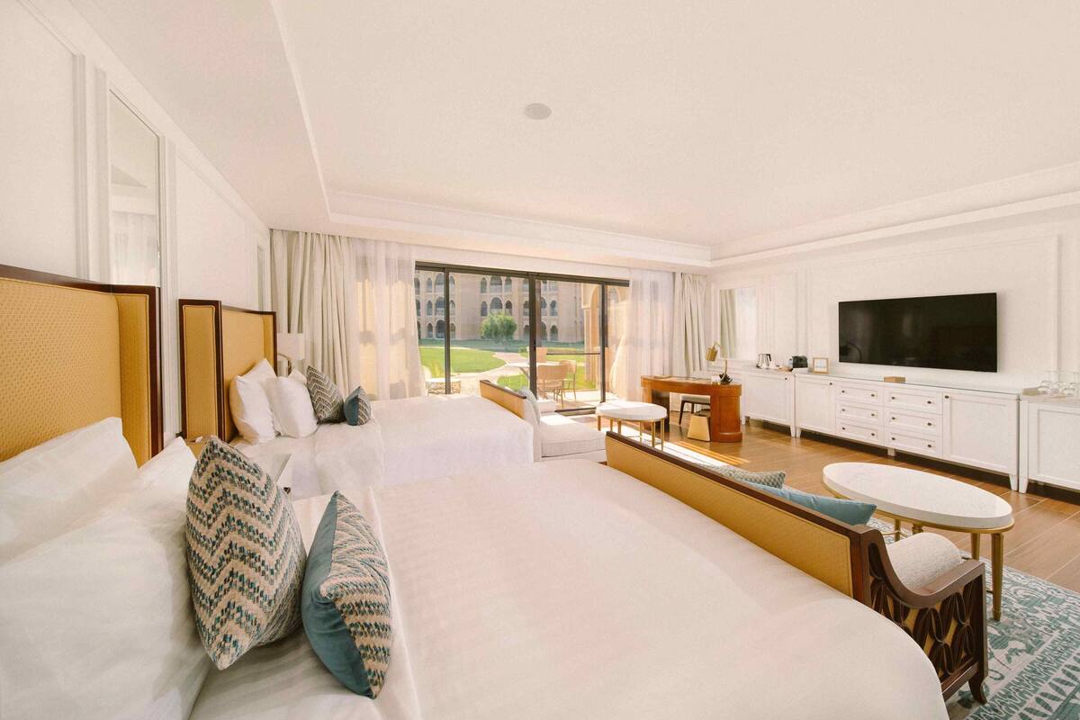 Quarto de hotel espaçoso com duas camas grandes, área de refeições, televisão e vista para um pátio.