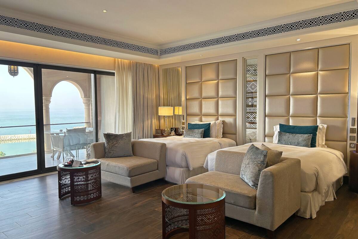 Quarto de hotel com uma cama grande, duas poltronas, uma mesa de centro de vidro e vista para o mar.