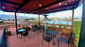 Um pátio externo com mesas e cadeiras, uma pérgola com luzes e a vista de um horizonte urbano com edifícios e montanhas ao fundo.
