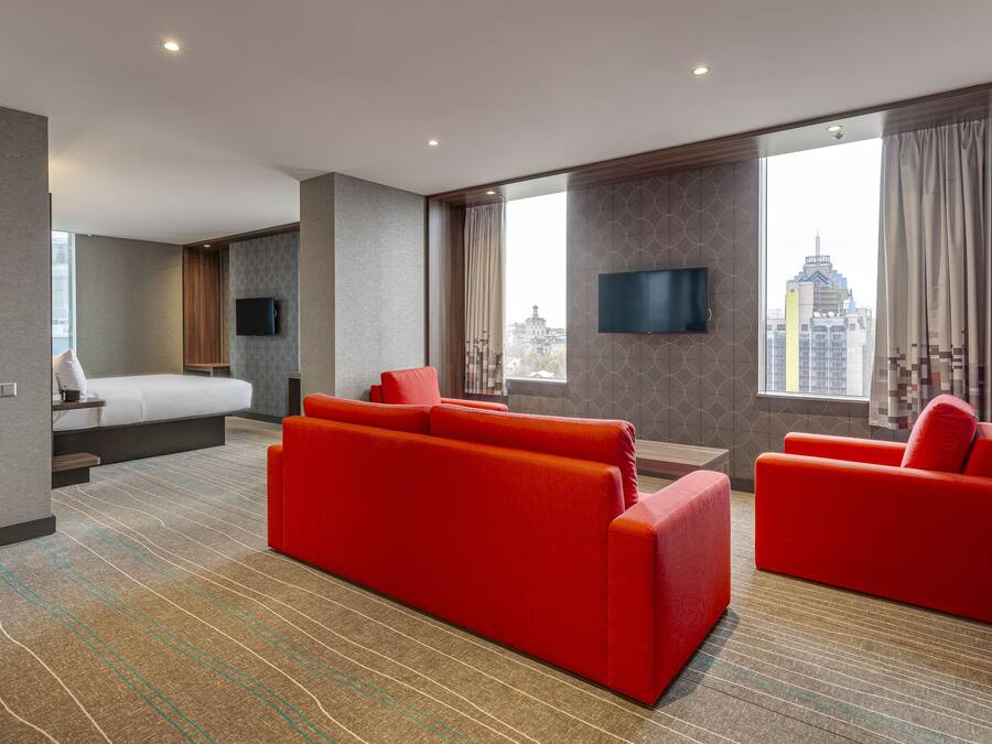 Quarto de hotel moderno com uma cama grande, um sofá vermelho, uma TV na parede e vista para a cidade pela janela.