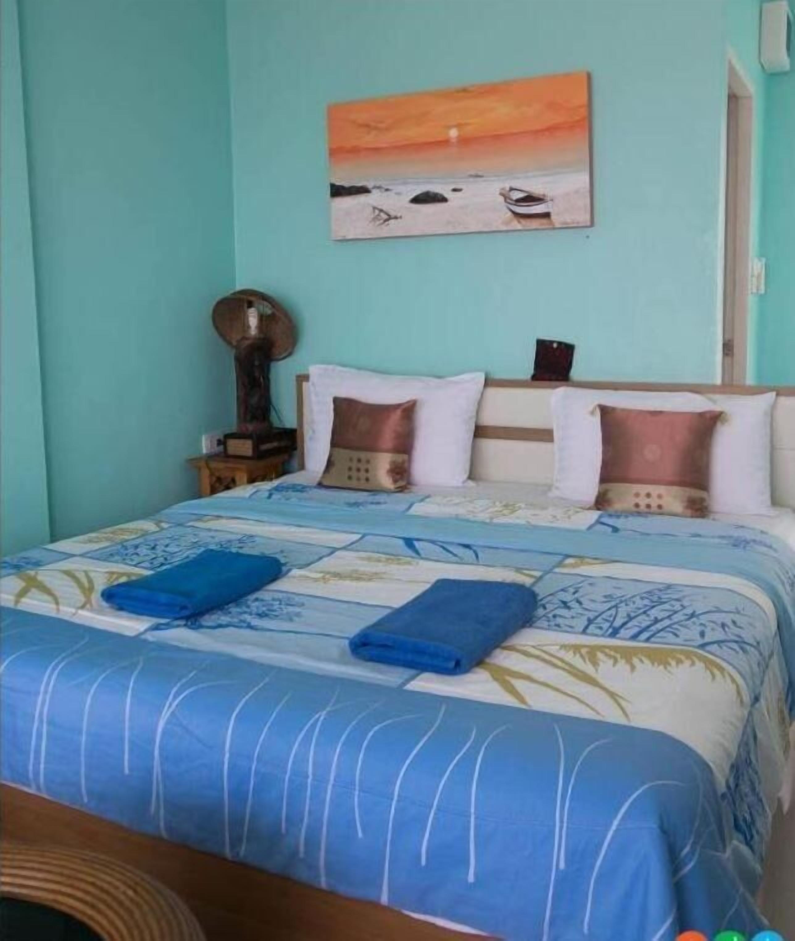Quarto com cama, almofadas, mesa de cabeceira, pintura na parede e ventilador.
