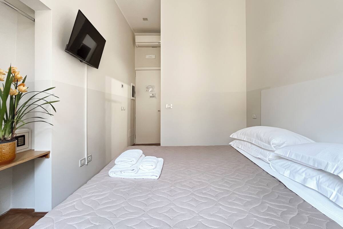 Quarto de hotel com cama, televisão presa na parede, planta em vaso e porta que dá para outro cômodo.