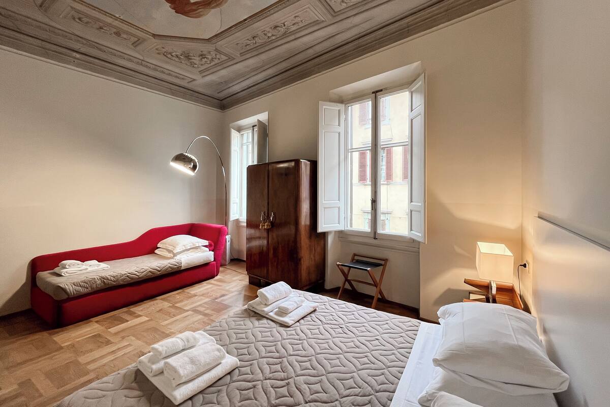 Quarto com um sofá vermelho, uma cama com colcha, um guarda-roupa de madeira e uma janela com persianas.
