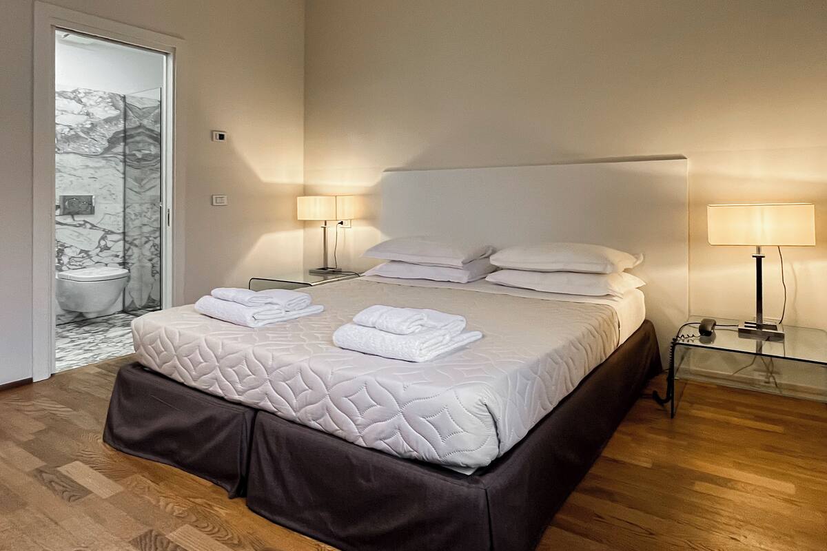 Cama bem arrumada com lençóis e almofadas brancas, duas luminárias de cabeceira e piso de madeira.