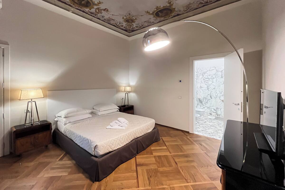 Quarto com cama, mesas de cabeceira, luminária e televisão.