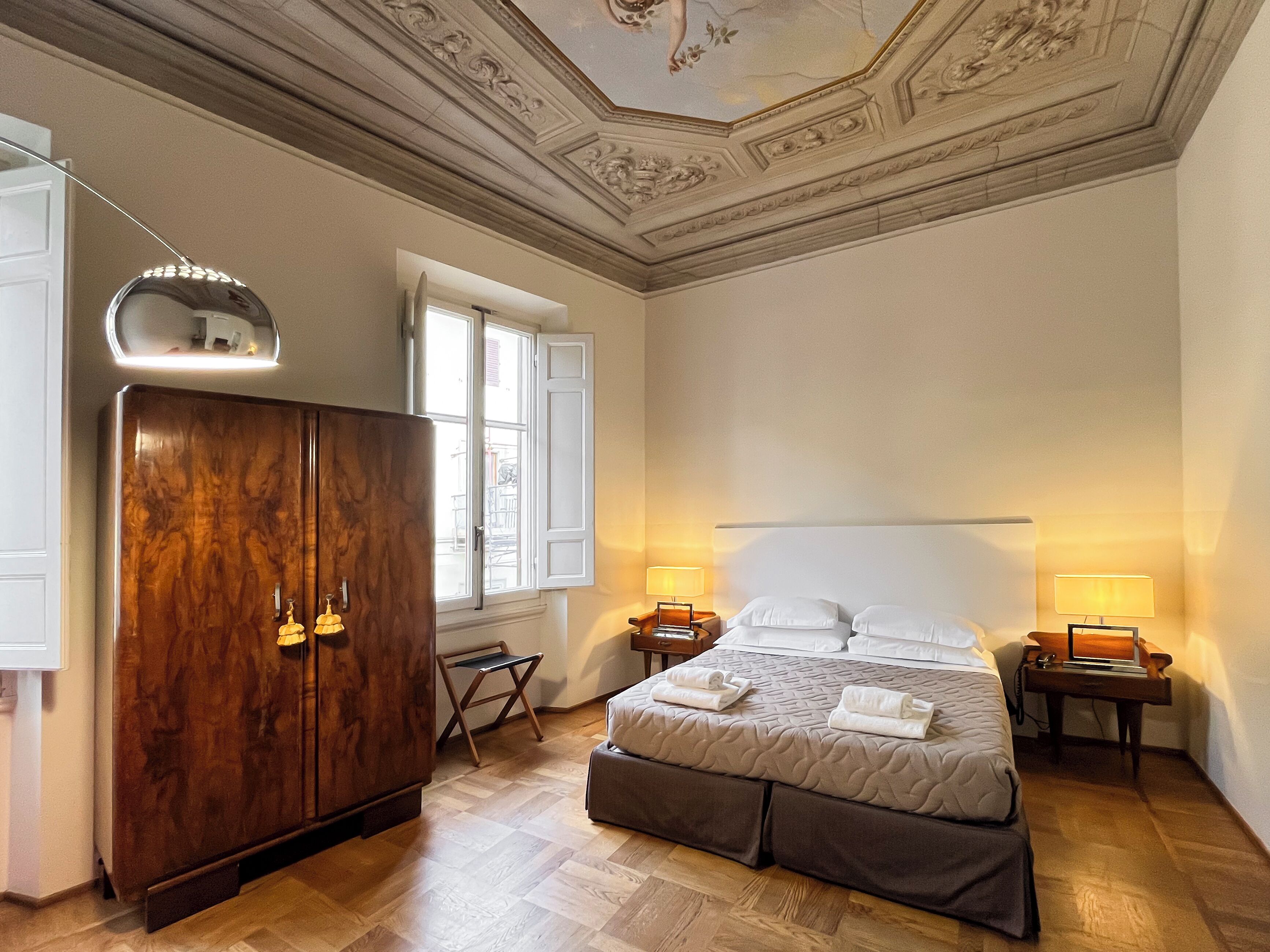 Quarto com guarda-roupa de madeira, cama com almofadas e cobertor, duas luminárias de cabeceira e uma janela com persianas.