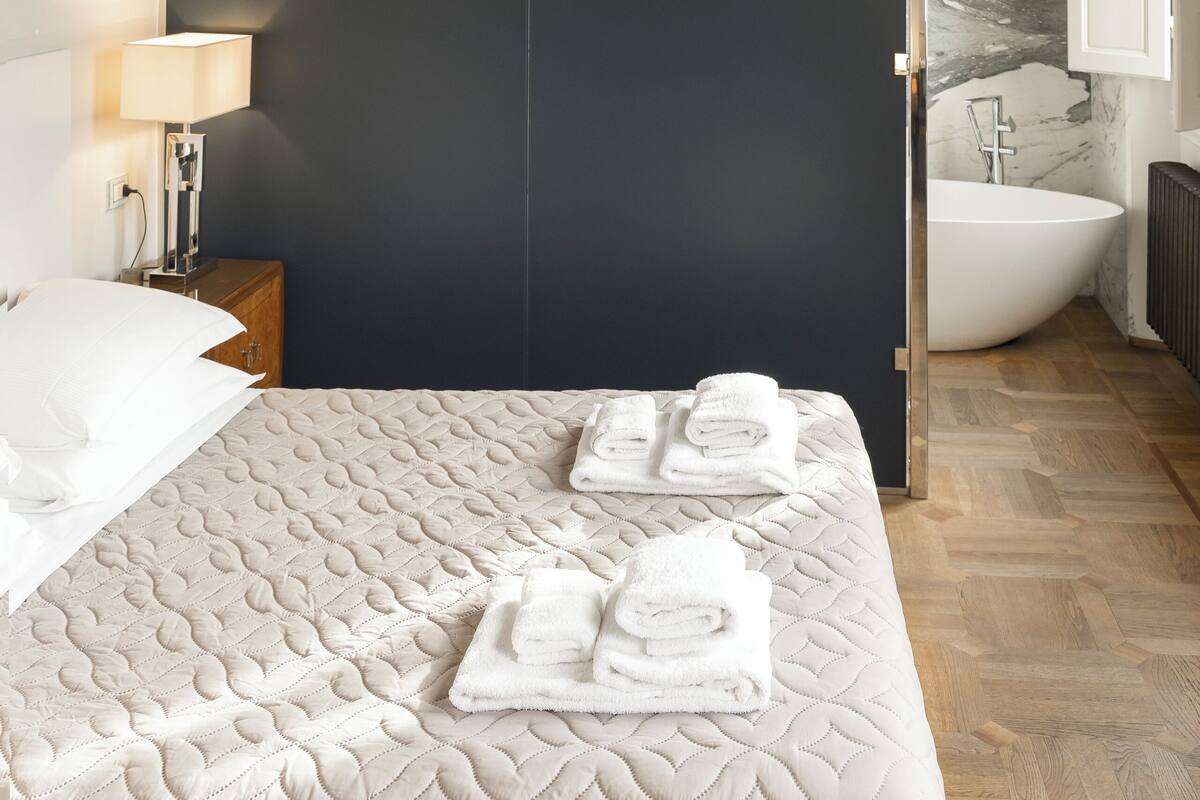 Quarto de hotel com cama, mesa de cabeceira, luminária, armário escuro, banheiro com banheira e parede de mármore.