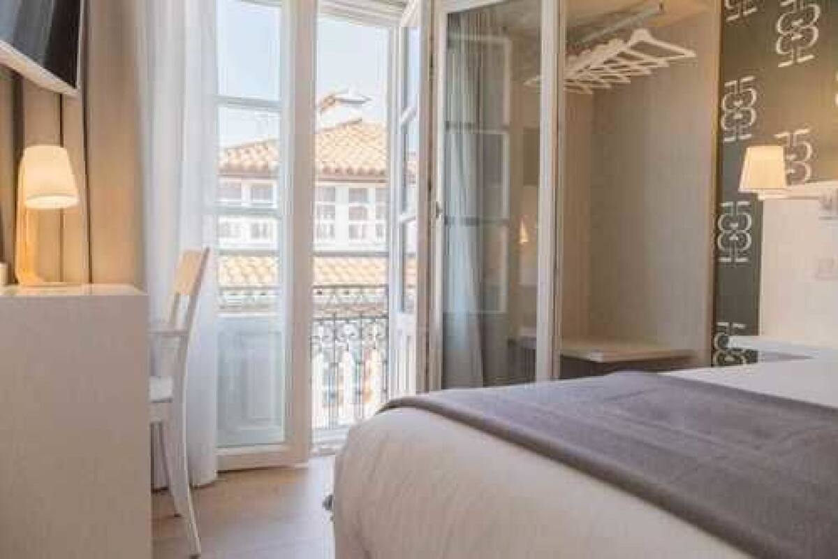 Quarto de hotel com cama, escrivaninha, cadeira, abajur e vista para o exterior.