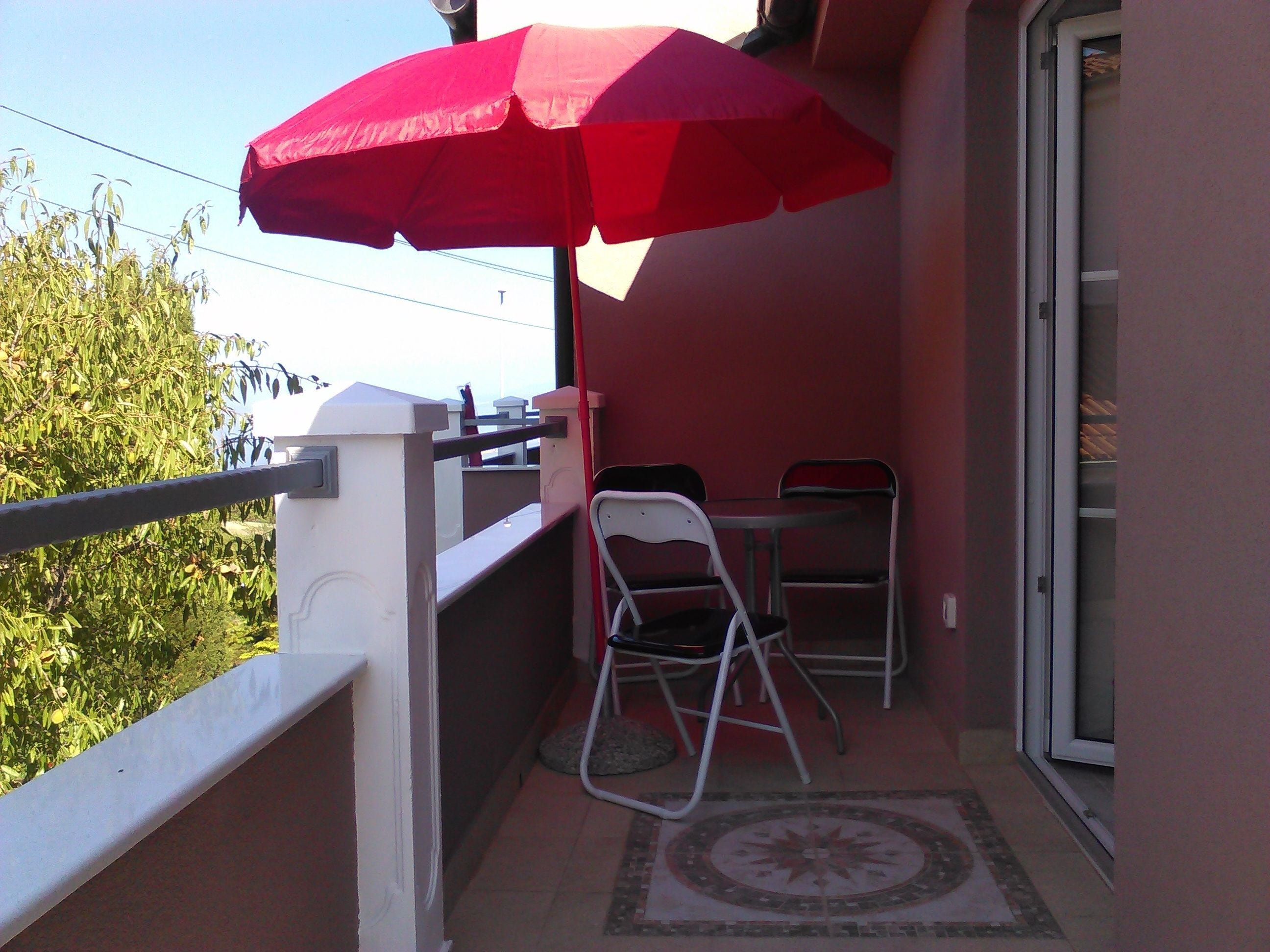 Uma varanda com um guarda-sol vermelho, duas cadeiras, uma mesa pequena e um tapete estampado.
