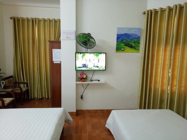 Quarto de hotel com duas camas, televisão na parede, um ventilador e um quadro de paisagem.