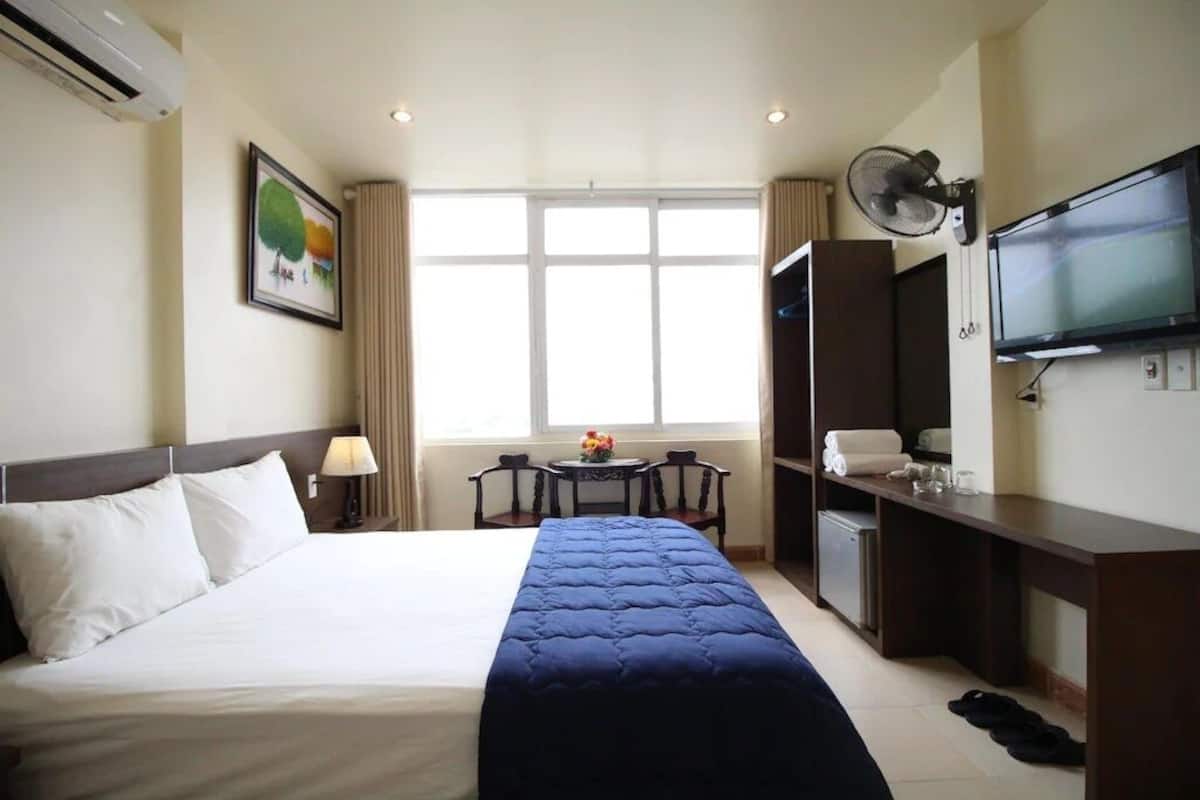 Quarto de hotel com cama, escrivaninha, cadeira, televisão e janela com cortinas.