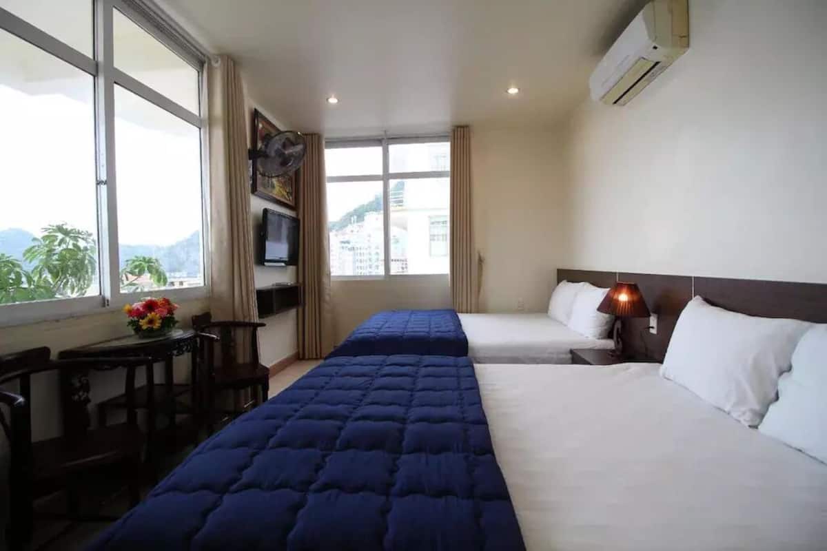 Quarto de hotel com duas camas, televisão, escrivaninha com cadeira e janela com vista para edifícios e montanhas.