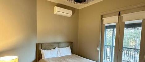 Um quarto com cama, ventilador de teto, ar condicionado, mesa de cabeceira e vista para o exterior através de uma porta de vidro.