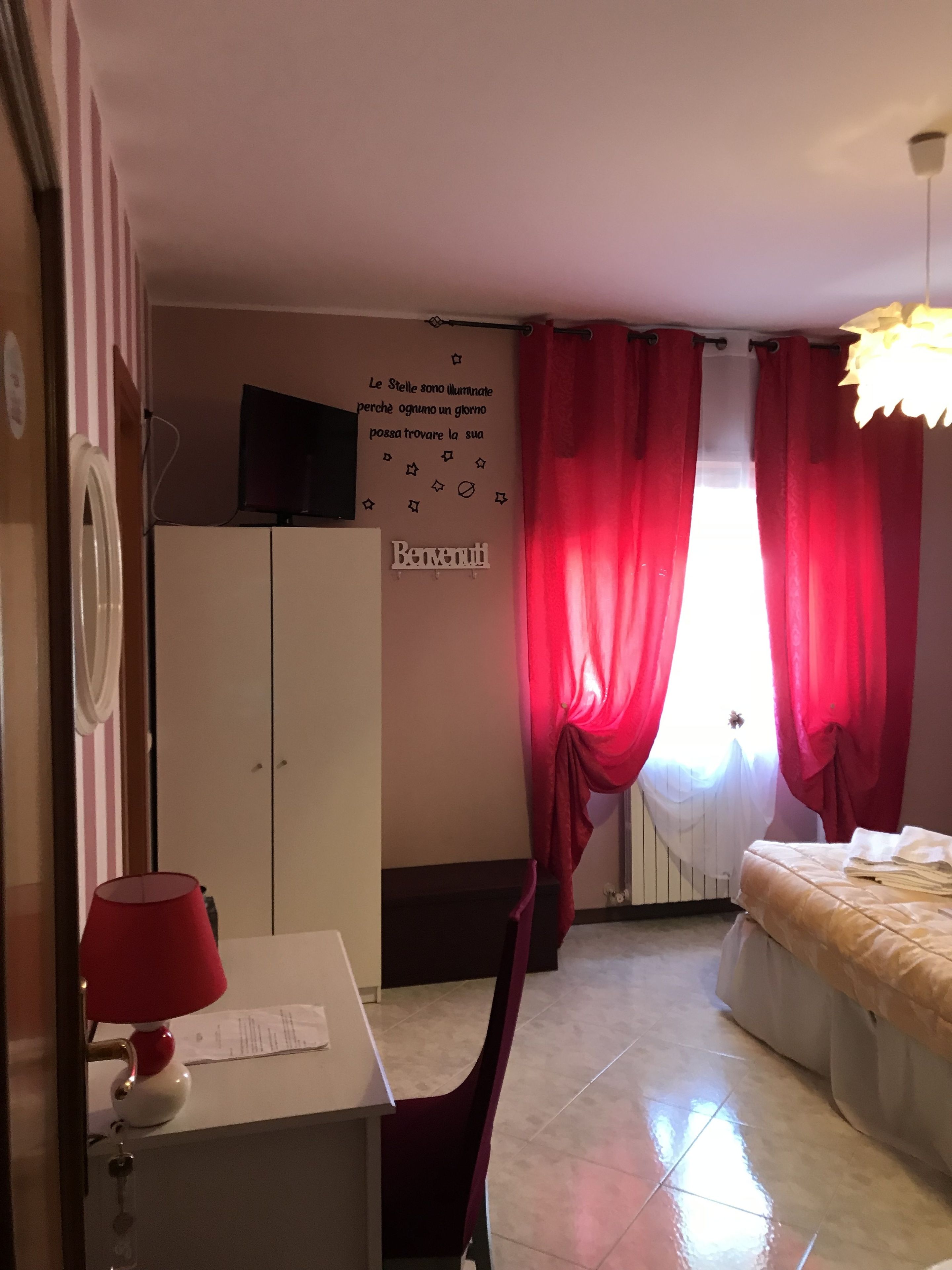 Quarto de hotel com cama, escrivaninha com luminária, guarda-roupa e uma placa na parede.