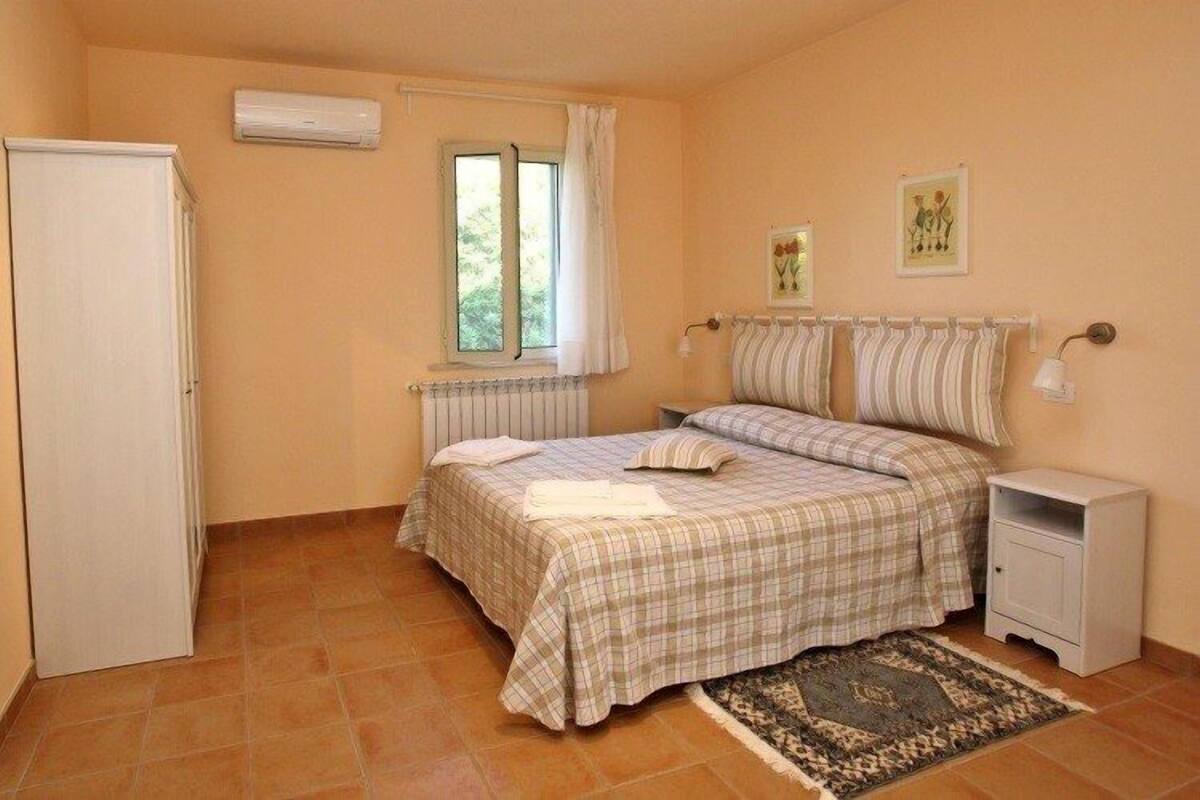 Quarto com cama, radiador, janela com cortinas, mesa de cabeceira e ar-condicionado fixado na parede.