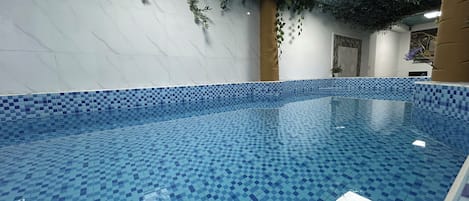 Piscina com azulejos azuis, rodeada por paredes com padrão azul e branco, e teto com folhagem artificial.