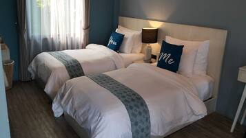 Quarto de hotel com duas camas, cada uma com roupa de cama branca e almofadas azuis. Há uma luminária na mesa de cabeceira, um cesto no chão e uma janela com cortinas finas.