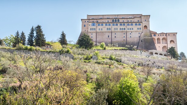 Um edifício grande e histórico com vários níveis e janelas em arco, rodeado por uma paisagem acidentada e vegetação escassa.