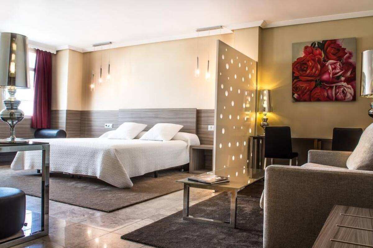 Quarto de hotel com cama, sofá, escrivaninha e cadeira. Há um quadro na parede e uma luminária na escrivaninha.