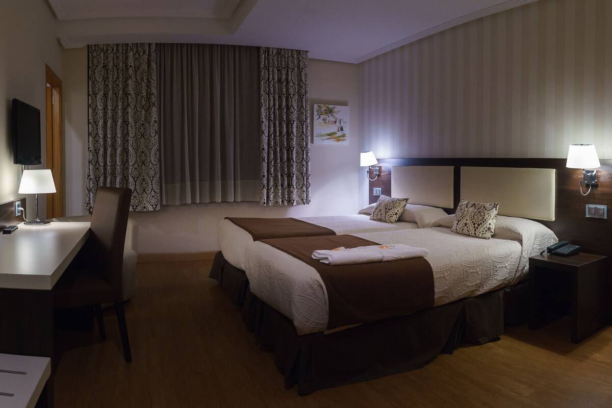 Quarto de hotel com cama, mesas de cabeceira, escrivaninha, cadeira e televisão.