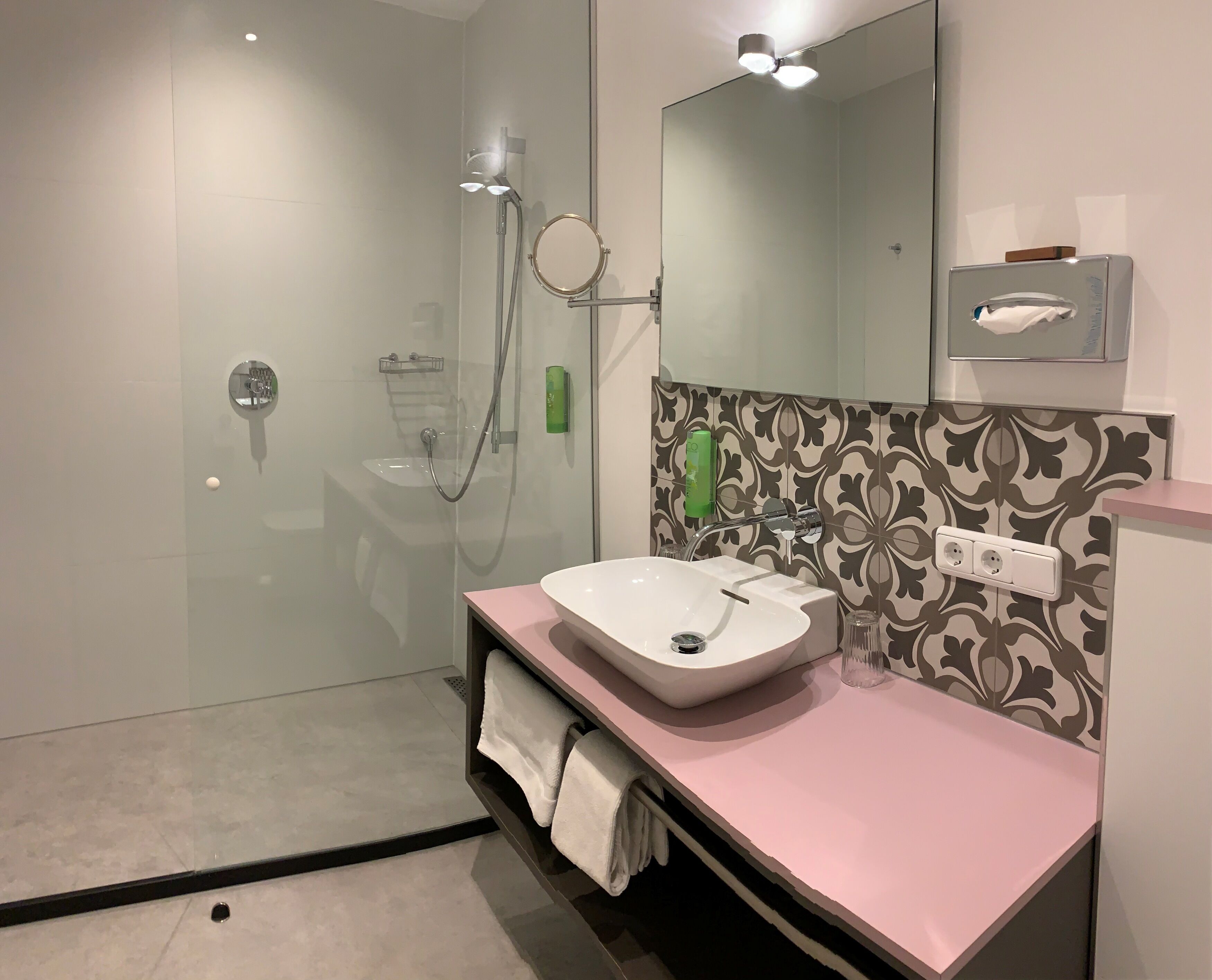 Casa de banho moderna com chuveiro em cabine de vidro, lavatório branco com bancada rosa e painel de azulejos estampados.