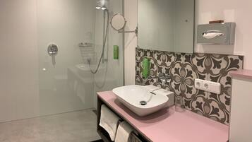 Casa de banho moderna com chuveiro em cabine de vidro, lavatório branco com bancada rosa e painel de azulejos estampados.
