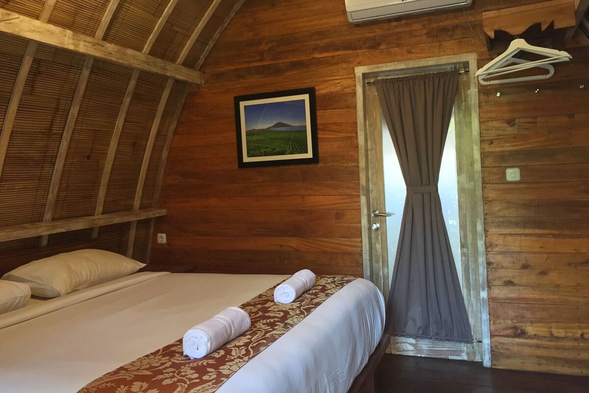 Um quarto de madeira com uma cama, um quadro, um ar condicionado e uma janela com cortinas.