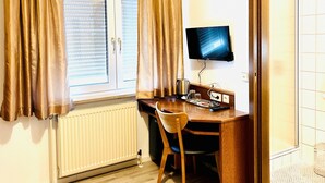 Quarto com escrivaninha de madeira, uma cadeira, televisão fixada na parede e janela com cortinas.