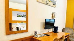 Uma escrivaninha de madeira com um monitor de computador, um controle remoto e um copo de água. Há uma cadeira e uma mesa pequena com uma revista e um copo.