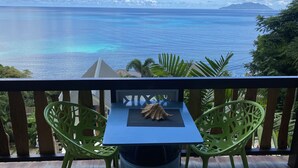 Varanda com mesa e cadeiras, vista para um mar azul e uma cordilheira distante.