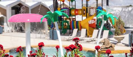 Área de piscina com um elemento aquático, estruturas de recreação coloridas e flores vermelhas em primeiro plano.