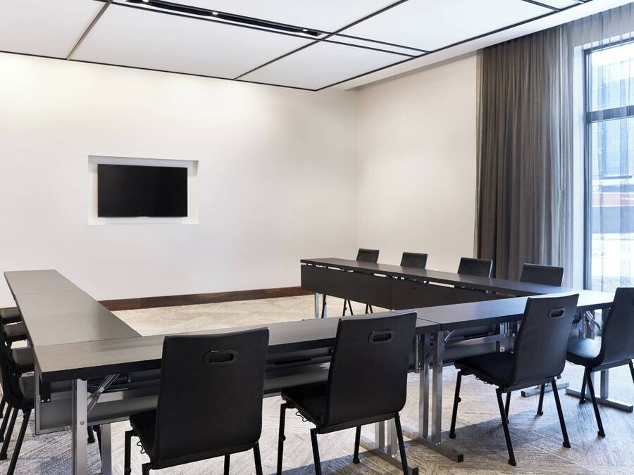 Uma sala de conferências com uma mesa comprida e cadeiras, uma TV de tela plana na parede e grandes janelas com cortinas.