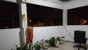 Uma varanda com plantas em vasos, uma cadeira e vista da cidade à noite.
