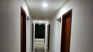 Um corredor longo e estreito com paredes brancas e chão de azulejos brilhantes. Há portas de madeira em ambos os lados e uma luminária no teto visível no final do corredor.