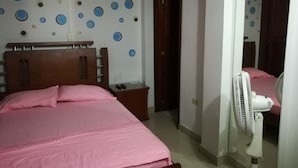 Quarto com uma cama, mesa de cabeceira, parede com padrões circulares, porta e espelho refletindo um ventilador.