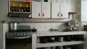 Cozinha com fogão, forno, panelas e utensílios diversos. Armários acima da bancada e um escorredor de louça à direita.