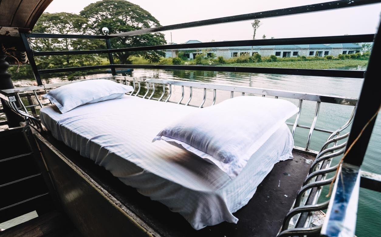 Uma cama com lençóis brancos numa varanda com guarda-corpo de metal, com vista para um corpo de água e um edifício ao fundo.